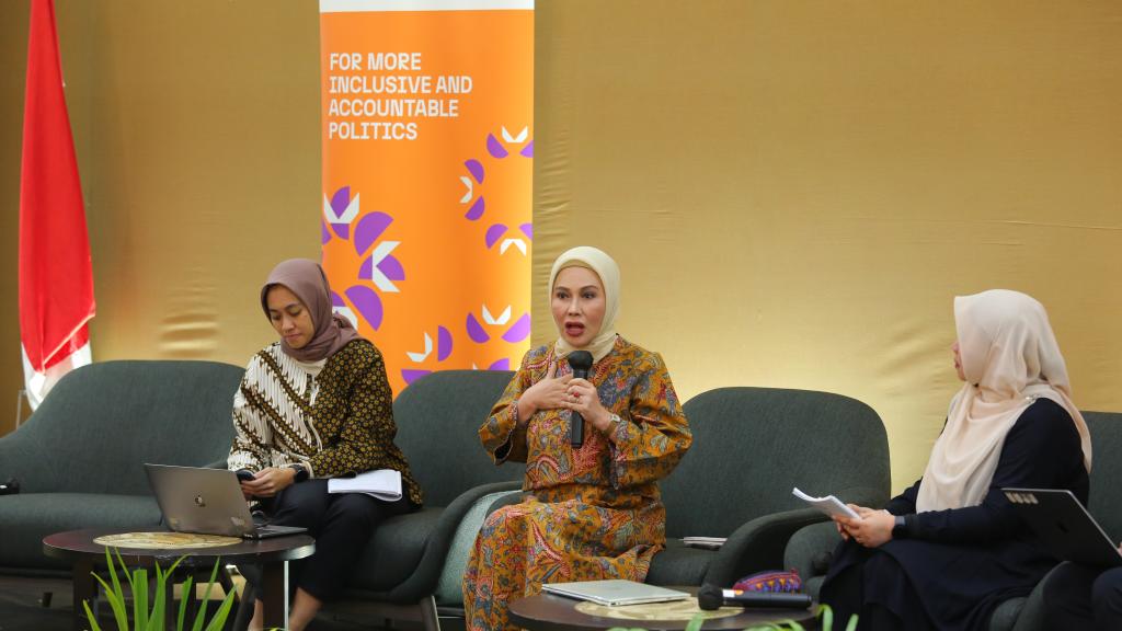 The image shows three women in hijabs sitting on a couch at a seminar. One woman is speaking into a hand-held mic. 